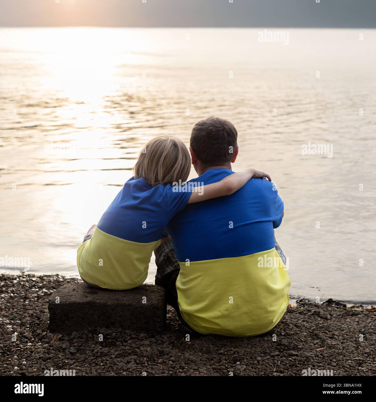 Dad son in identical yellow and blue T-shirts sit hugging side by side ...