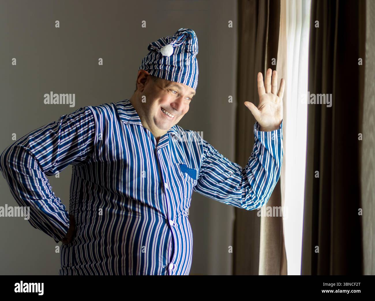 Cheerful fat man wearing striped pajamas stands by a window and waving ...