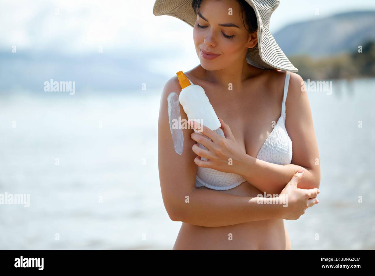 A beautiful woman stands on a sunny beach, applying sunscreen to ...