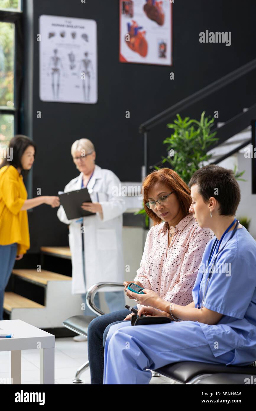 Nurse assisting asian patient in measuring her sugar levels with glucometer for a routine ...