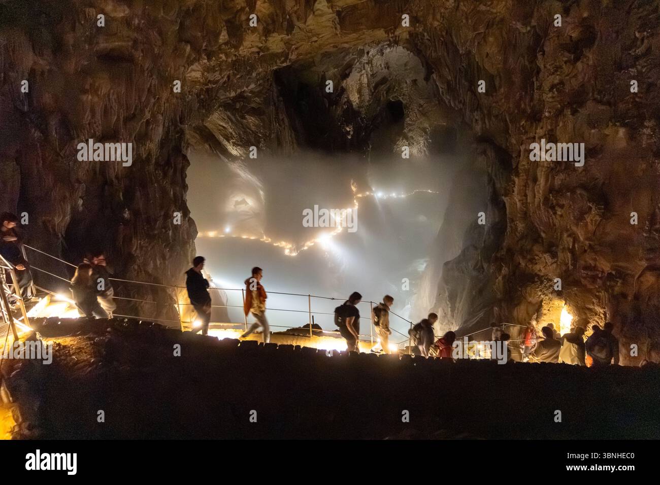 Tourists exploring a massive cave, walking on an illuminated footpath ...