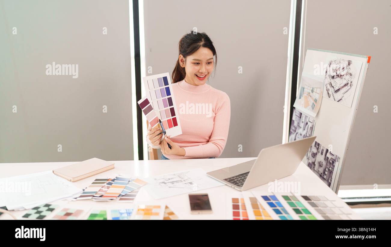 A young Asian woman interior designer at her desk selects a color ...