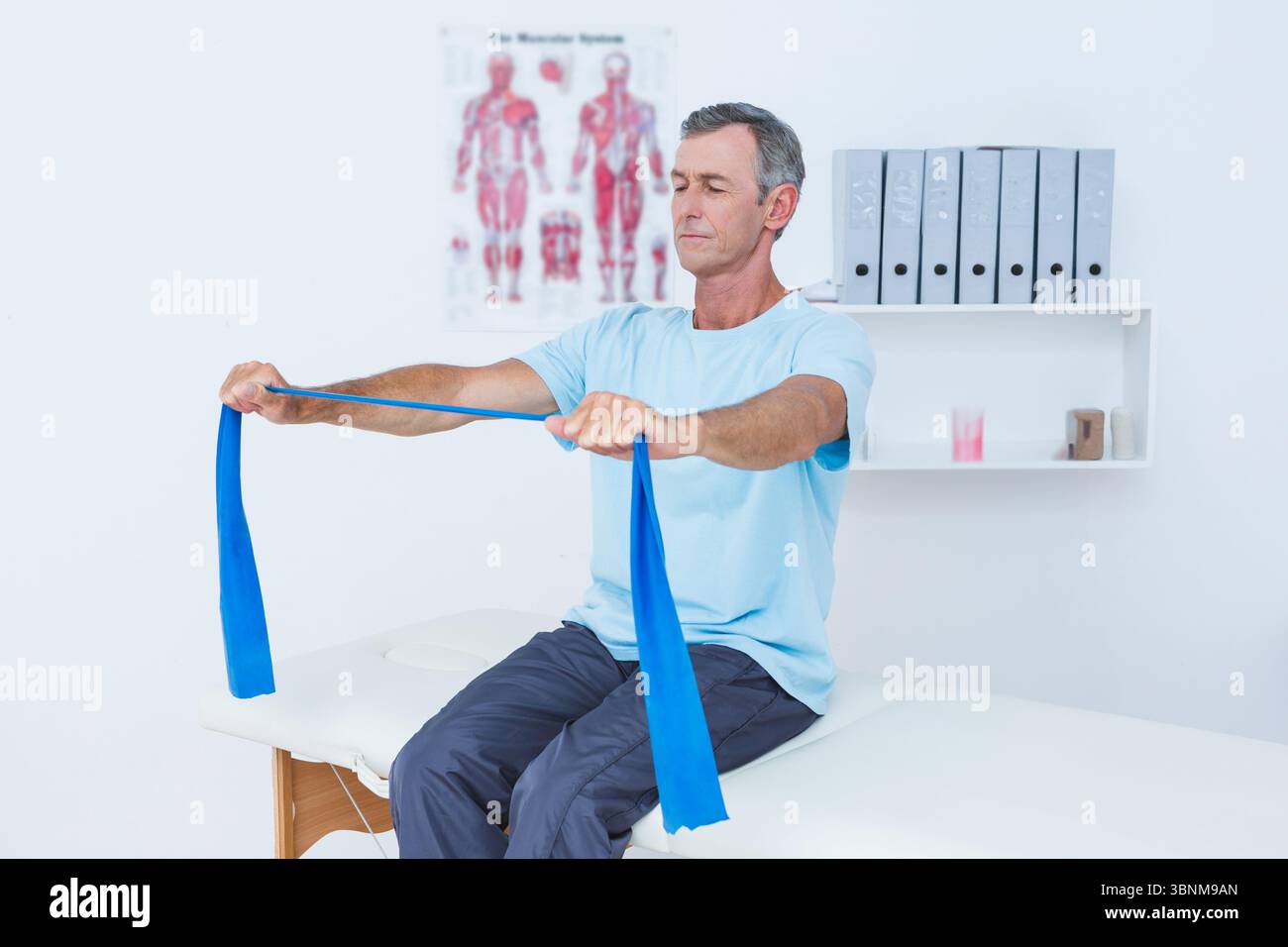 Middle-aged man pulling blue elastic band on table in therapy room with ...