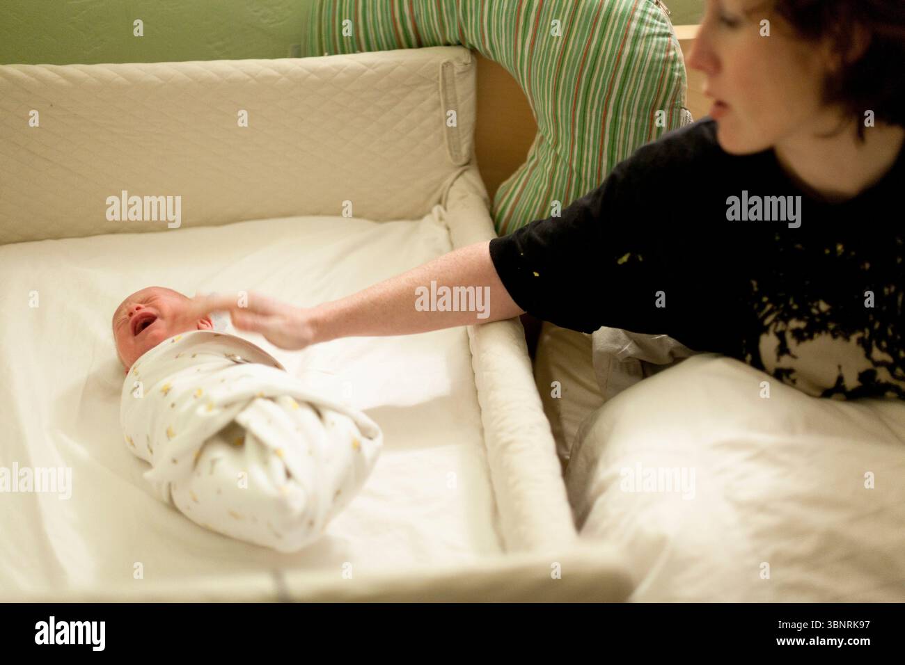 A woman soothes a swaddled crying baby in a co-sleeper crib next to her ...
