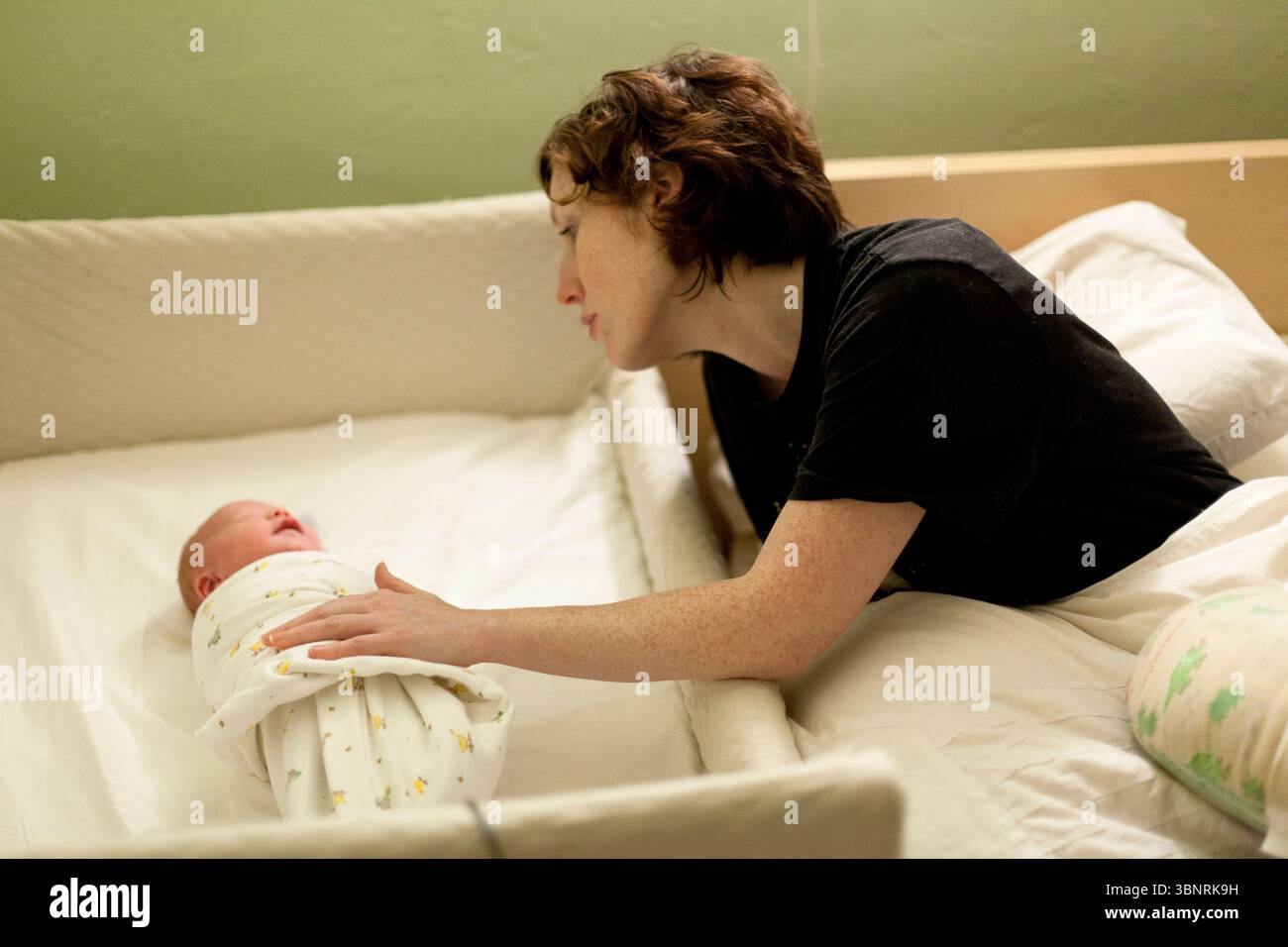 A woman soothes a swaddled crying baby in a co-sleeper crib next to her ...