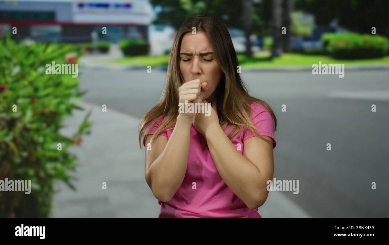 Woman coughing while holding her throat, wearing pink shirt outdoors on ...