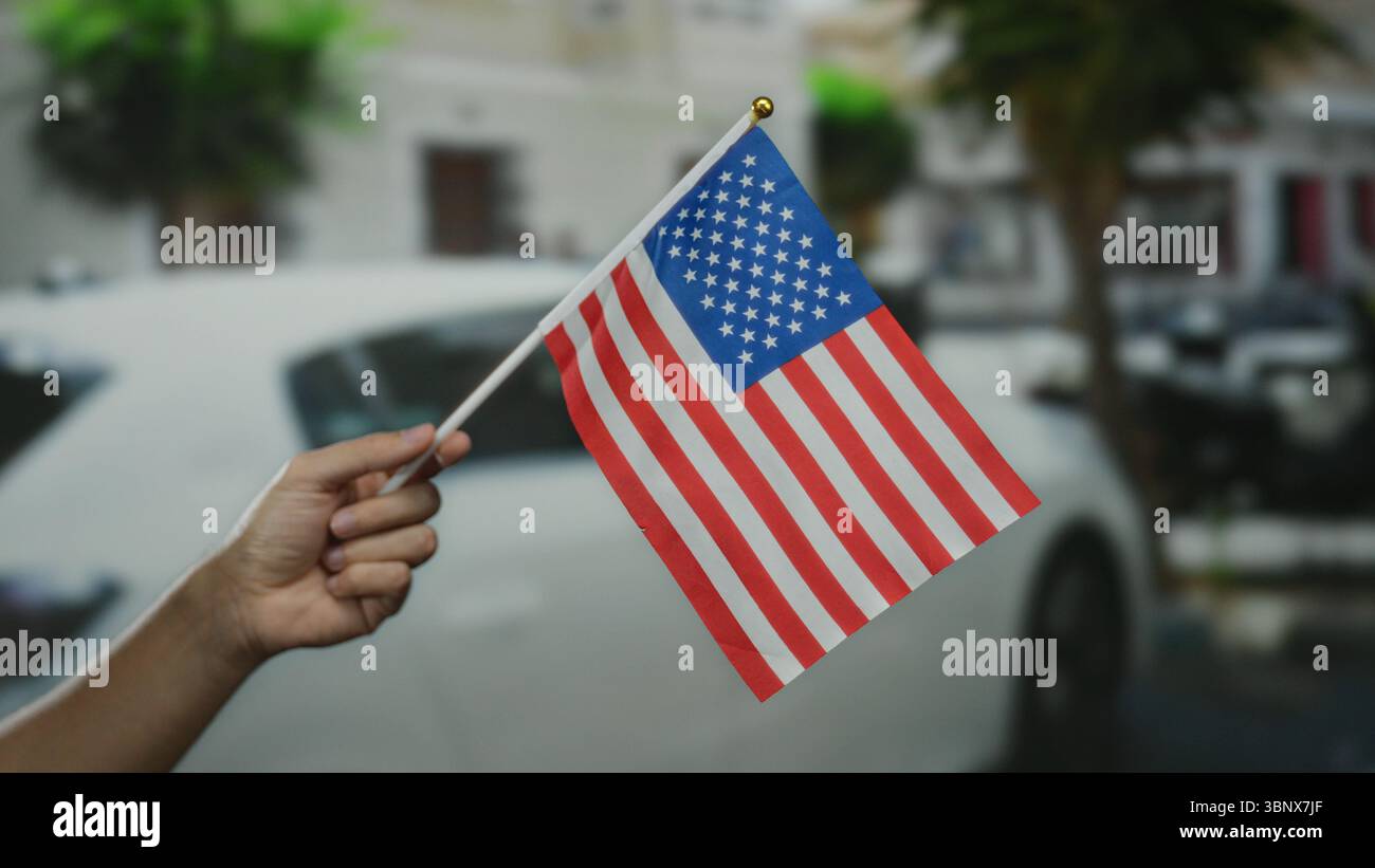 Man hand holding american flag in city street scene with cars and trees ...