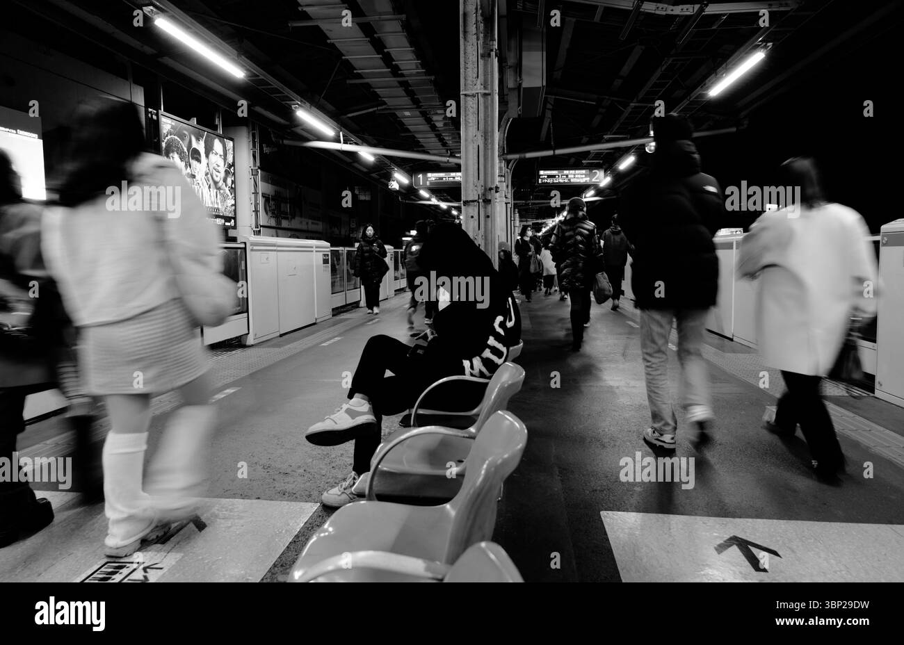 Black-and-white metro platform at night, hooded figure seated, blurred ...