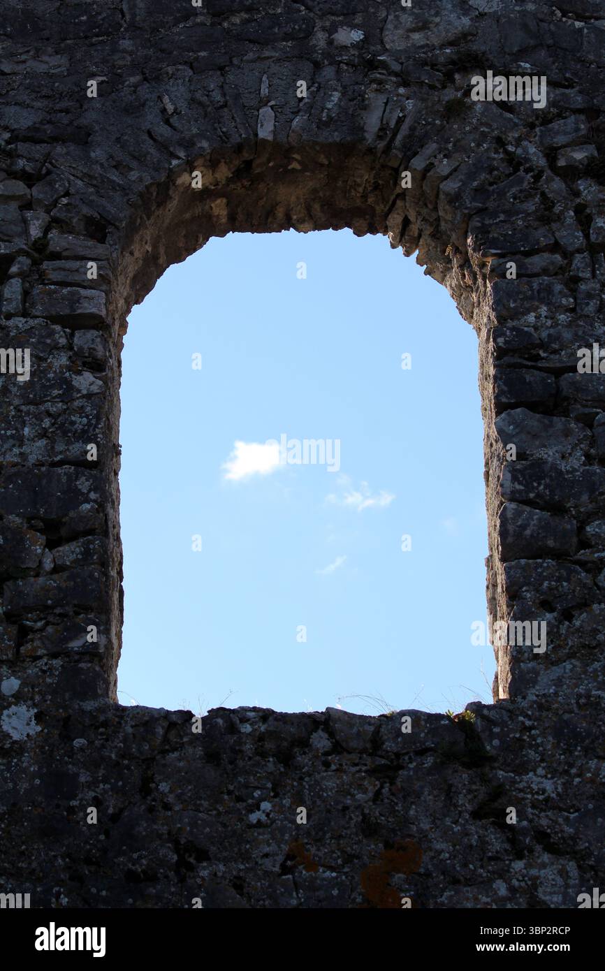 A rough-hewn stone window arch in ancient ruins frames a patch of blue ...