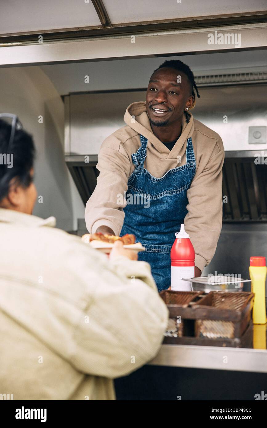 Smiling young man serving hot dog to customer while standing in food ...