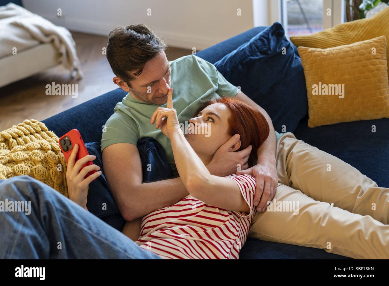 Couple in love lying on sofa with smartphone and hugging while bonding at home. Young family ...