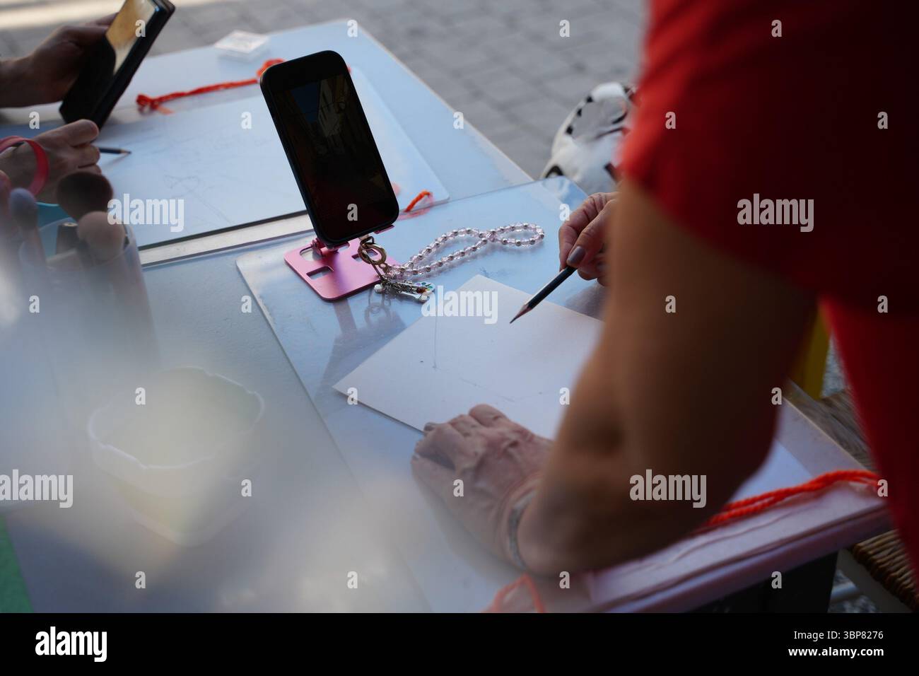 An artist seated at an outdoor table sketches on paper using a pencil ...