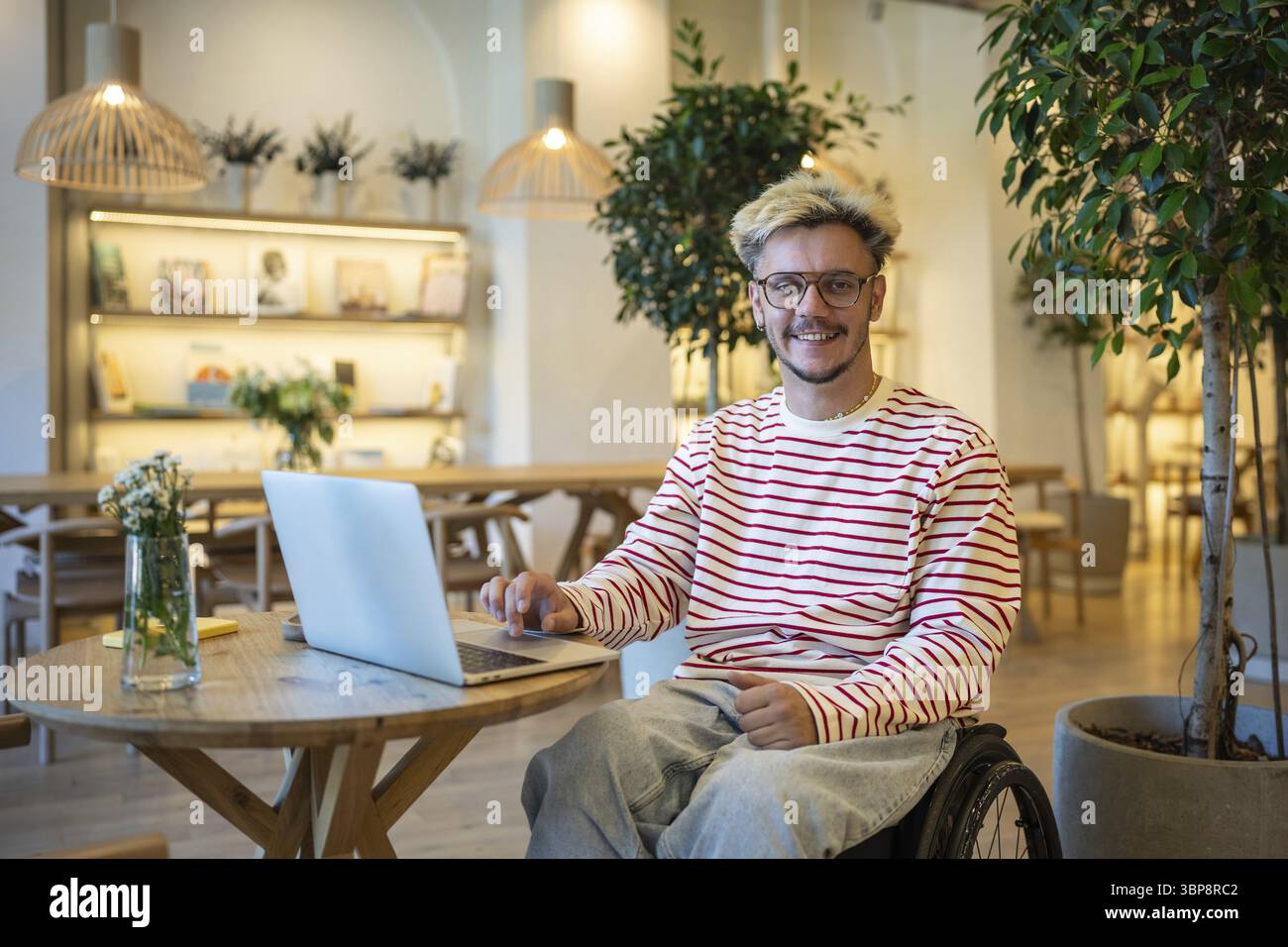 Happy, content disabled man in wheelchair works on laptop in cafe. Digital nomad handles remote ...