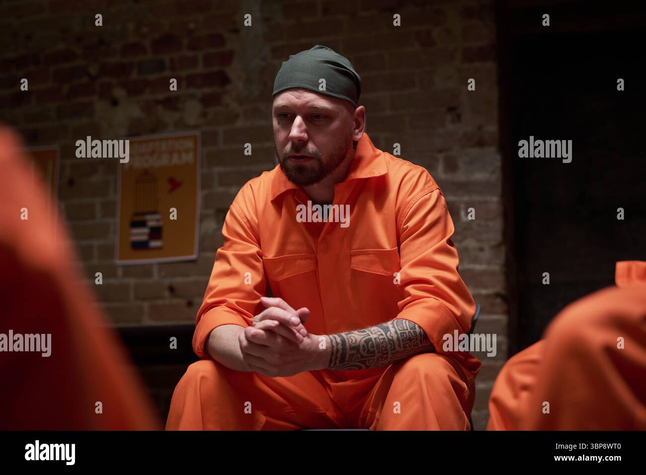 Caucasian man with beard and tattooed forearm sitting in orange prison ...