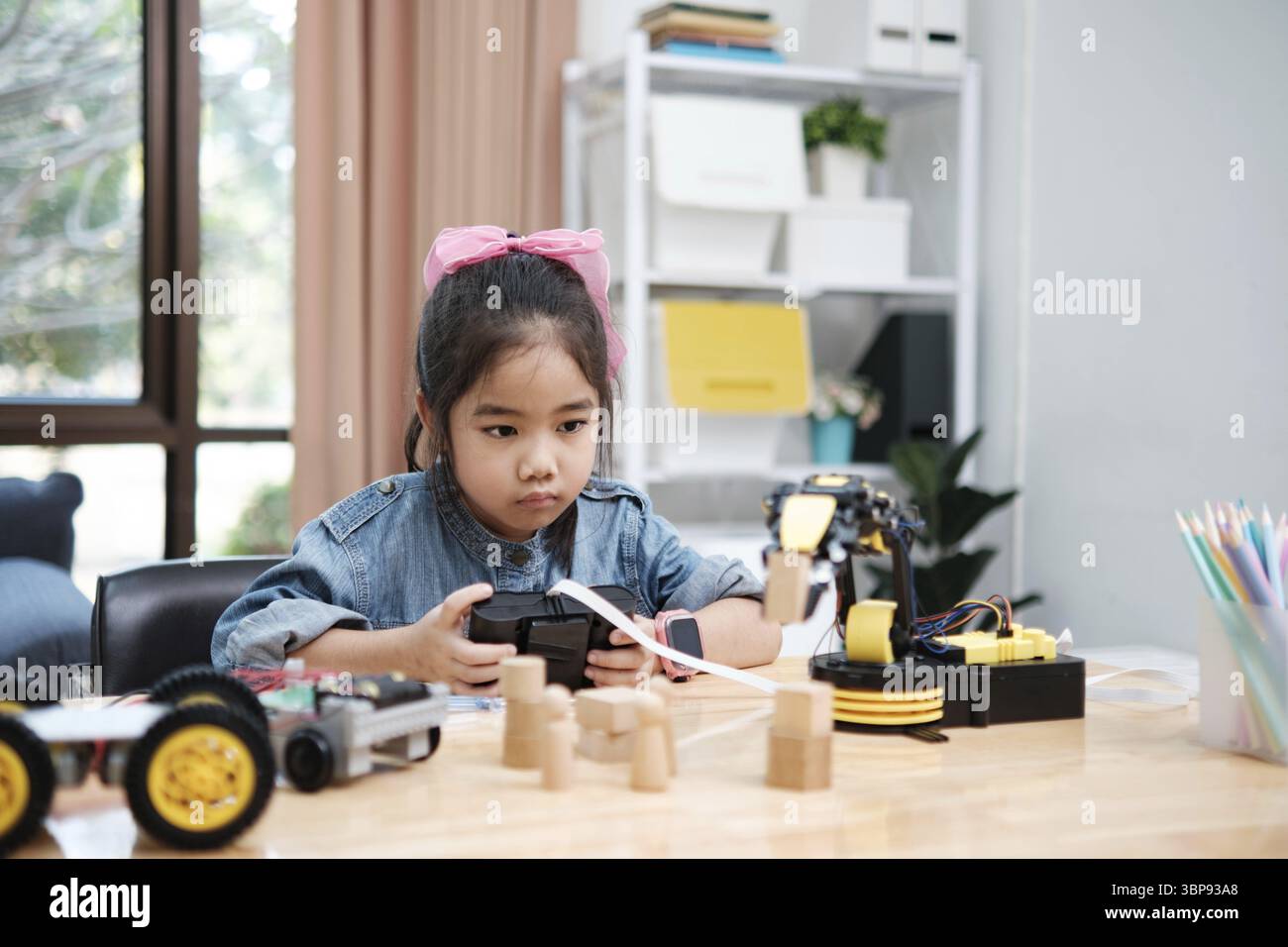 A primary school girl focuses on operating a robotic arm with a remote control, demonstrating ...