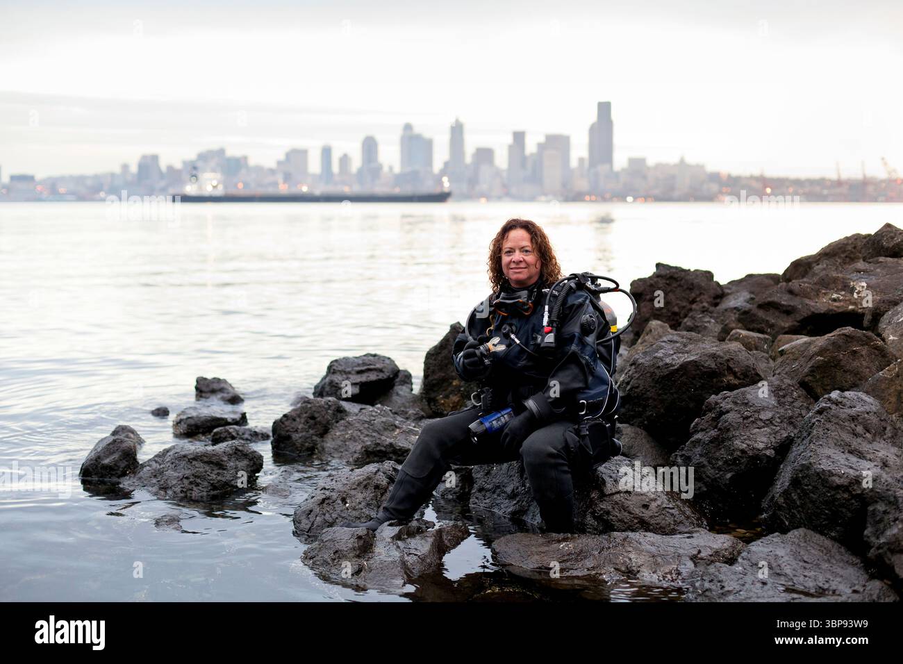A female scuba diver sits on a rock at the water edge with the city ...