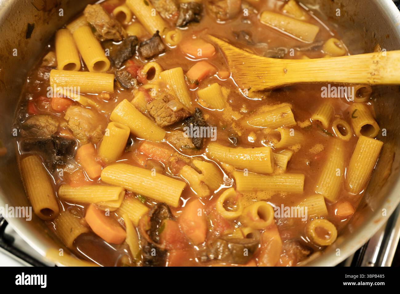 Wooden spoon stirring a hearty beef stew with rigatoni pasta, mushrooms ...