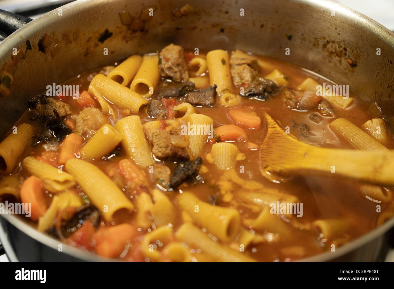 Wooden spoon stirring hearty beef stew with rigatoni pasta, mushrooms ...