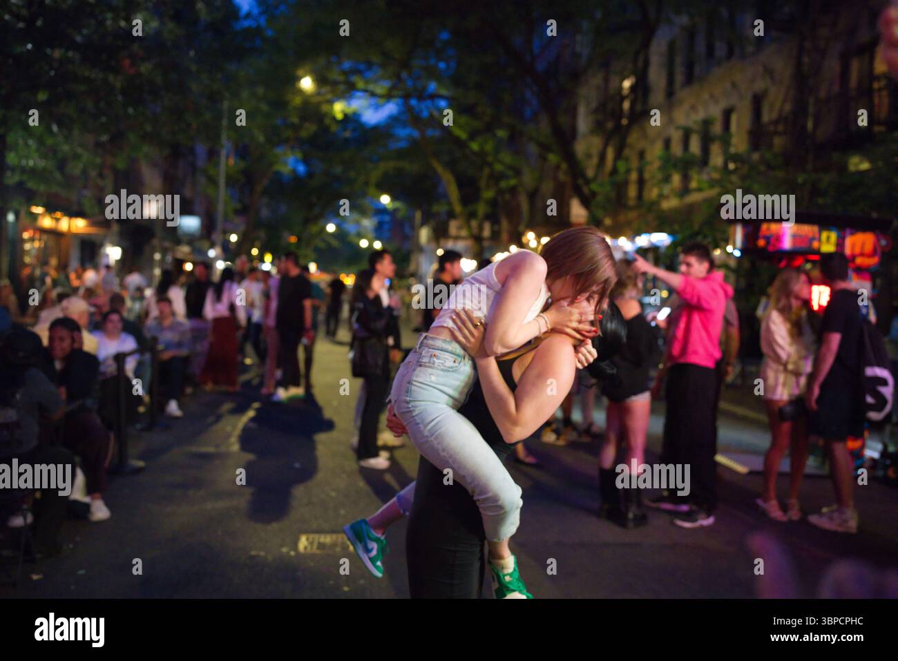 Two women embrace and kiss on a city street at night, one lifting the ...