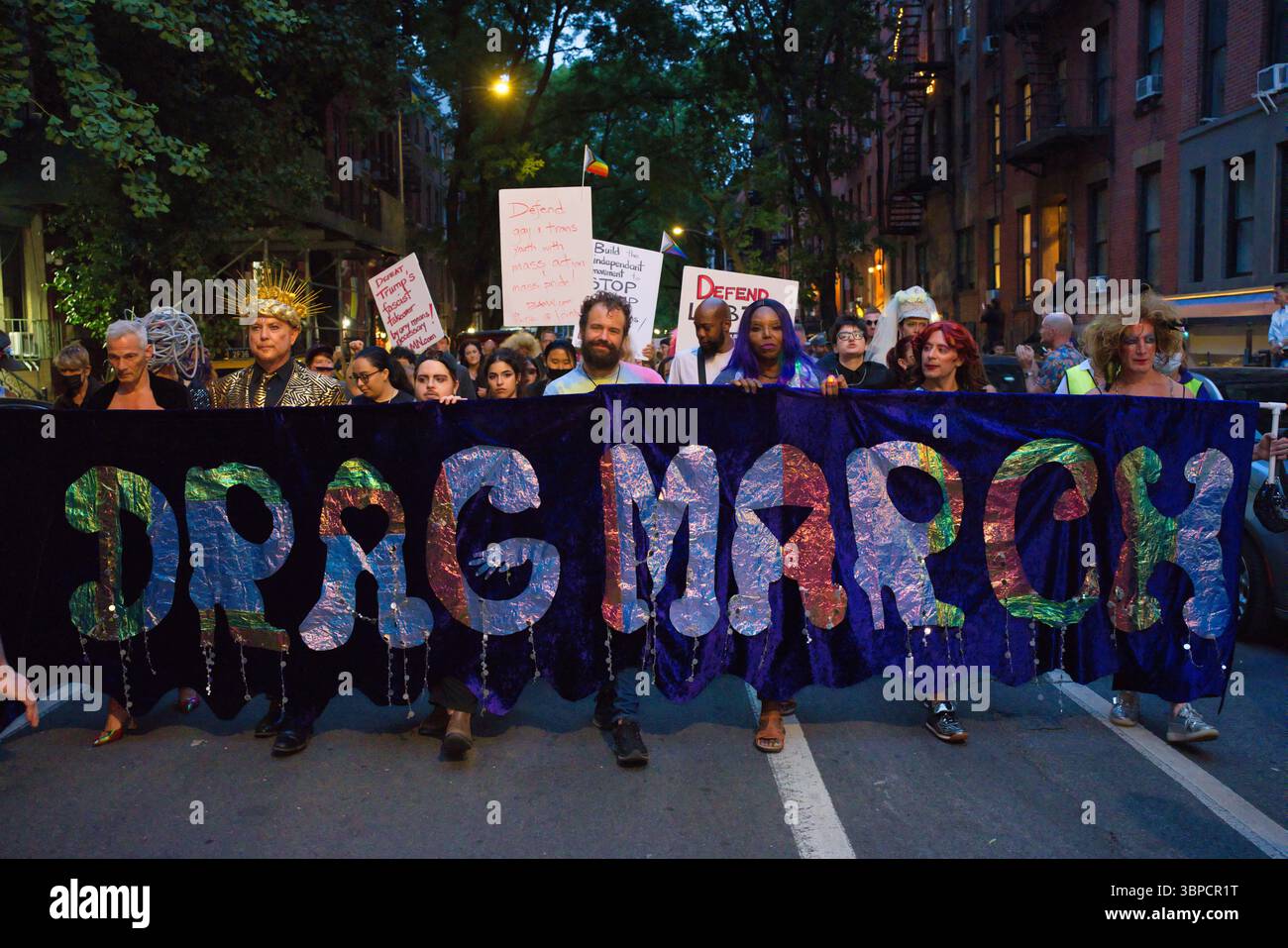 A group of people, many in drag, hold a large banner reading "DRAG ...