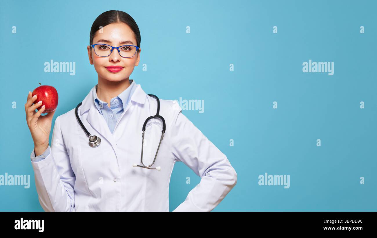 Portrait of a young female doctor in a lab coat holding a red apple ...