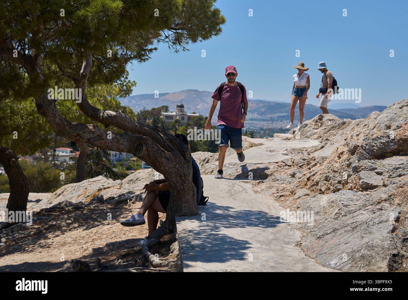A woman sits in the shade of a tree as other tourists walk on ...