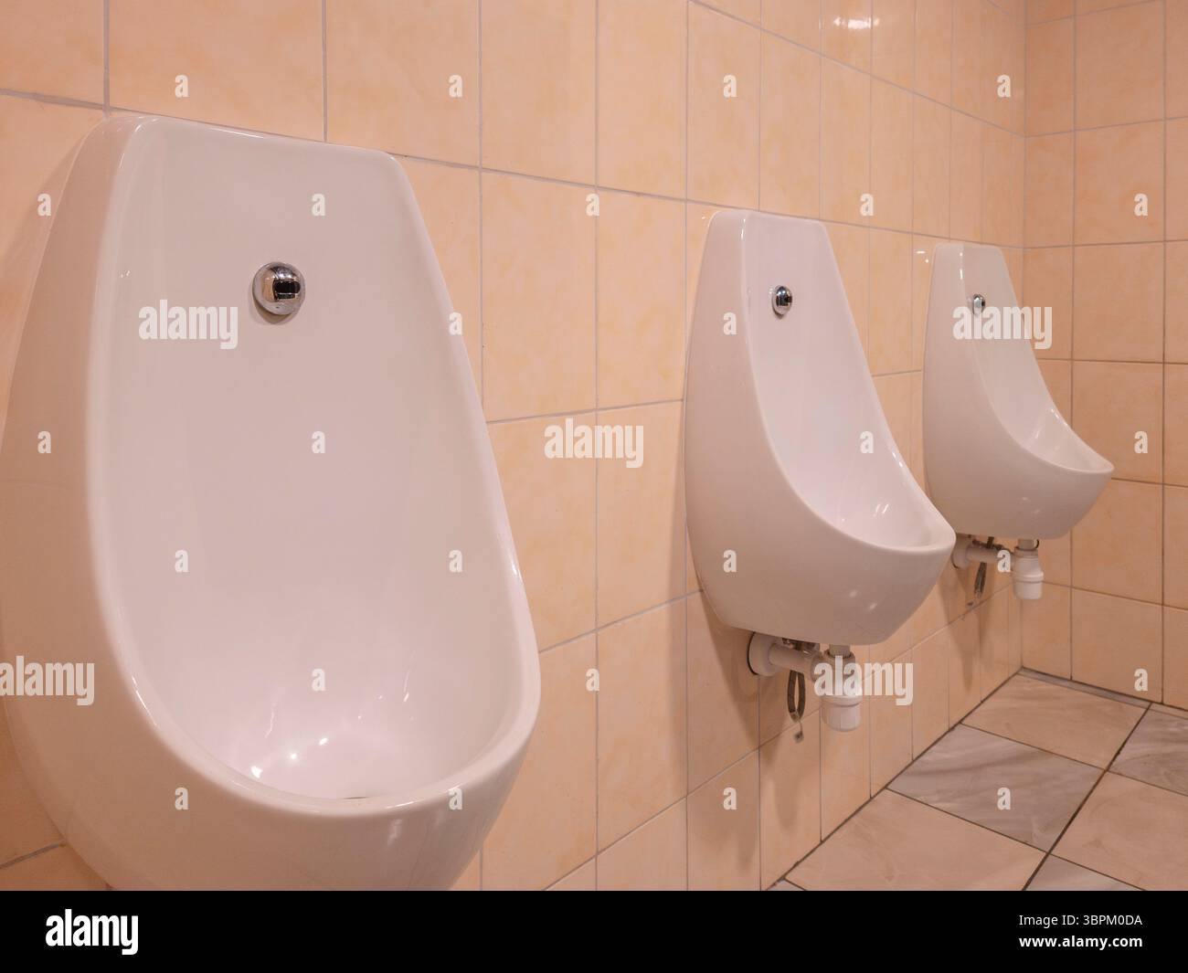 Three white urinals are lined up against a light-colored tiled wall in ...