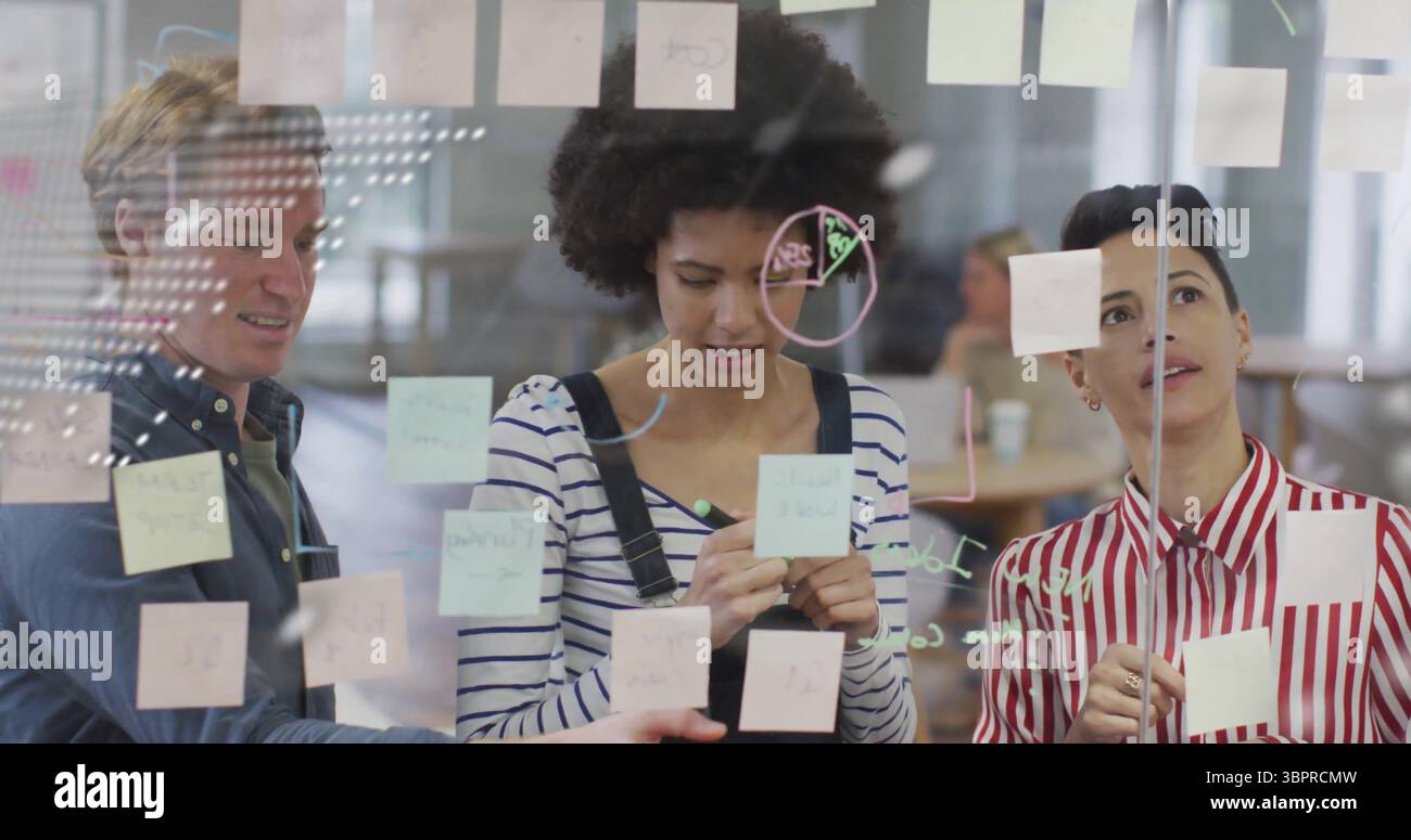 Collaborating team placing pastel sticky notes on glass wall in office ...