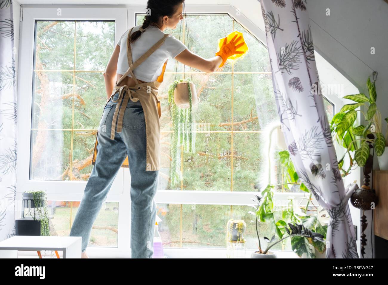 Woman in rubber gloves and apron manually washes attic window of house with rag cleaner and mop ...