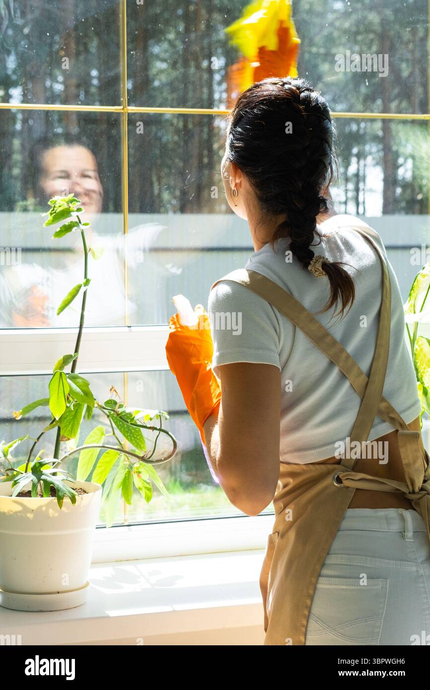 Woman in rubber gloves and apron manually washes window of house with rag cleaner and mop inside ...