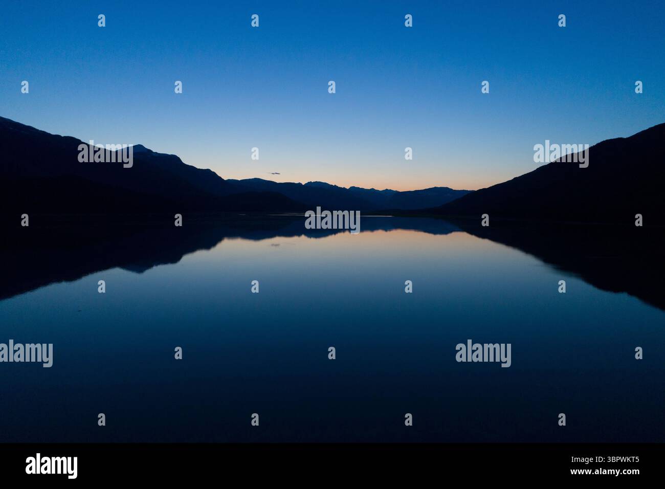 Silhouetted peaks are casting reflection on lake water and dusky sky is ...
