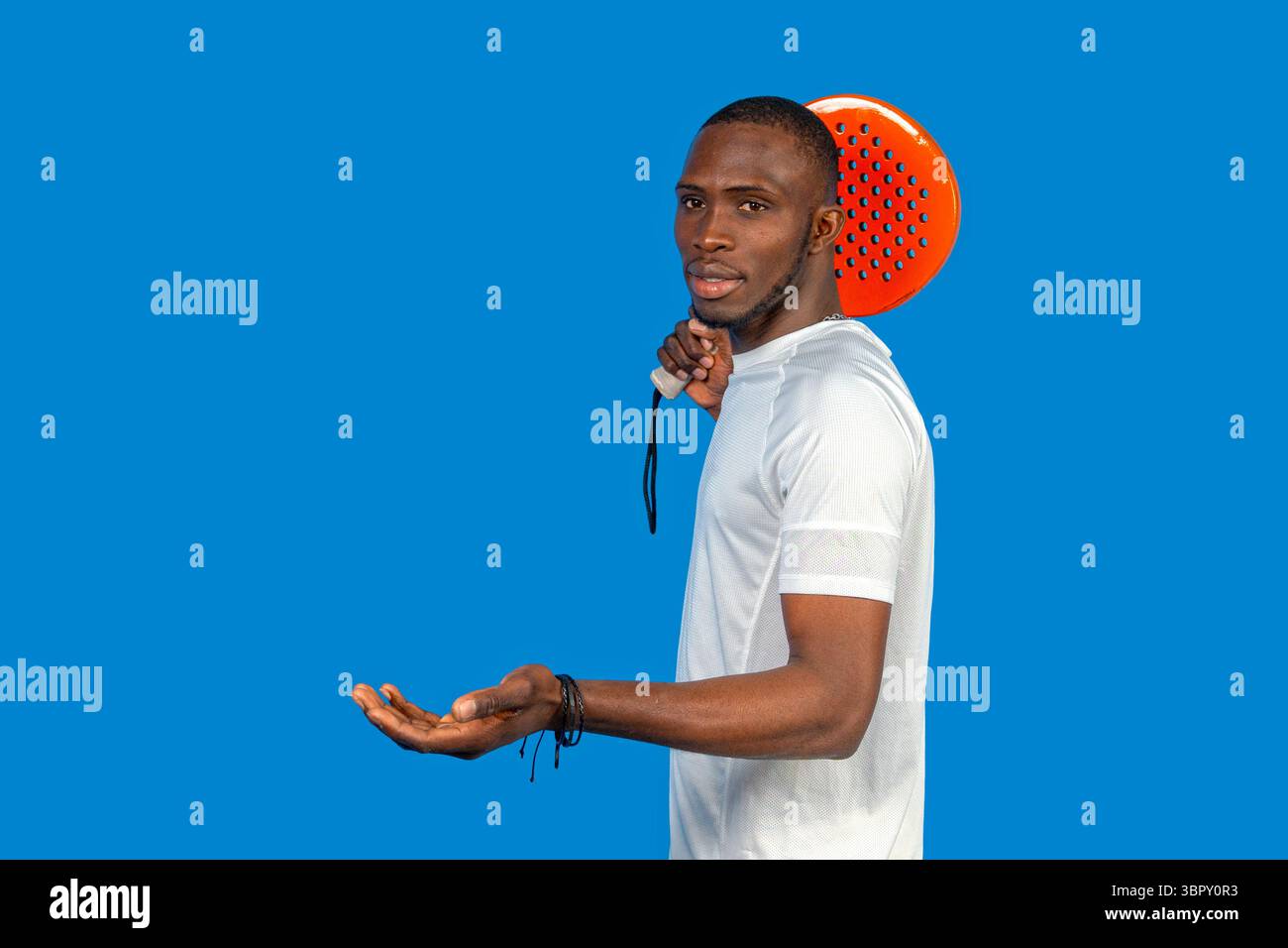 Young black man holding a padel racket showing his open hand on a blue ...