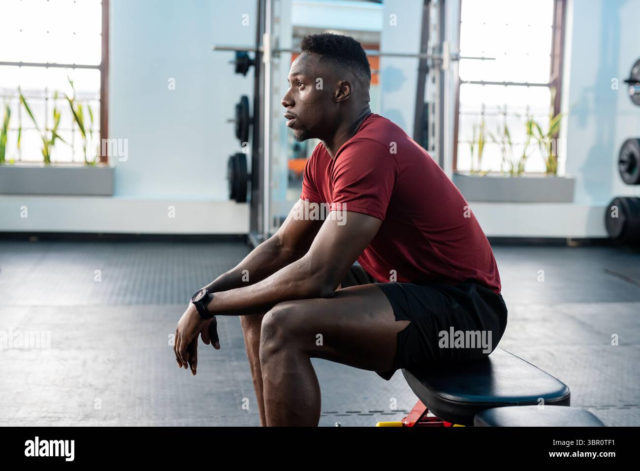 African American man resting on weight bench in gym wearing red athletic T-shirt, smartwatch ...