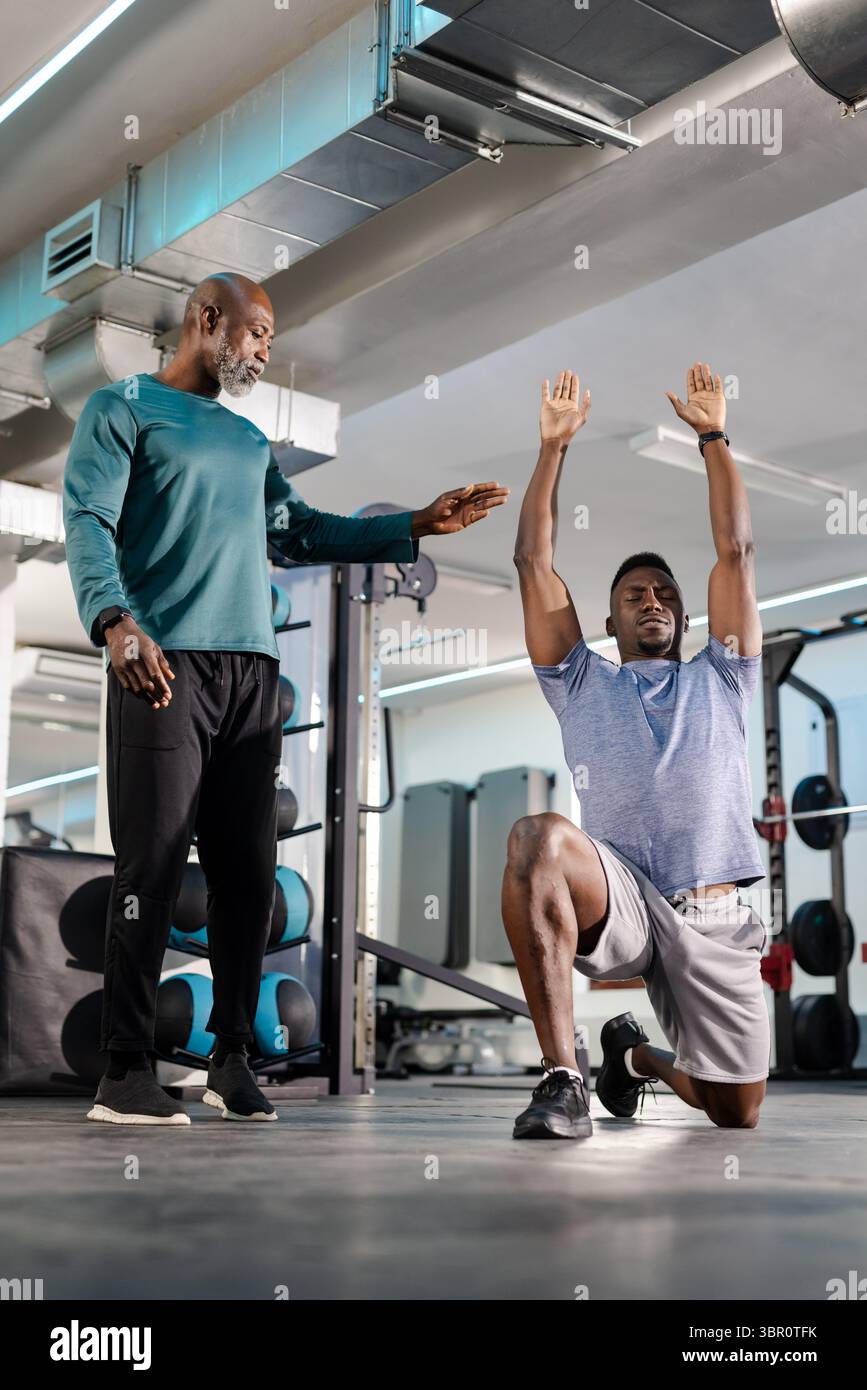 Senior African American male trainer guiding client doing forward lunge at gym with kettlebell ...