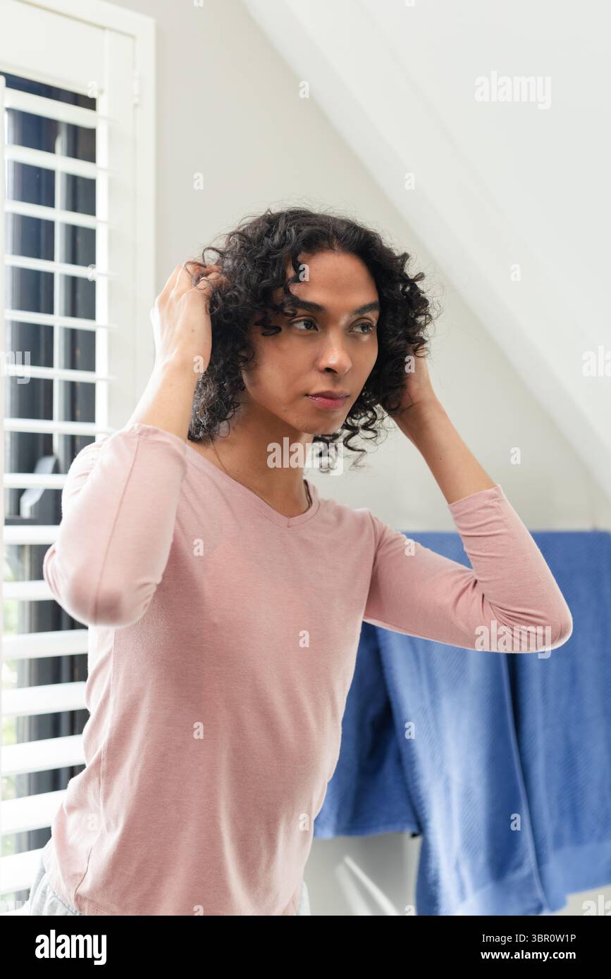 Non-binary person adjusting curly hair in bathroom near window with ...