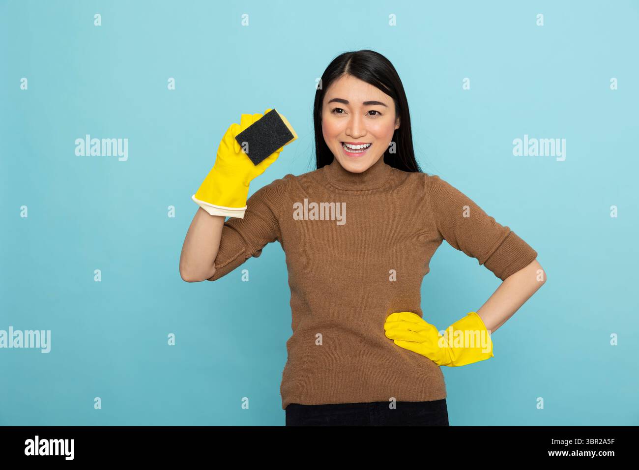 Smiling asian housemaid stands with hand on waist and another holding ...