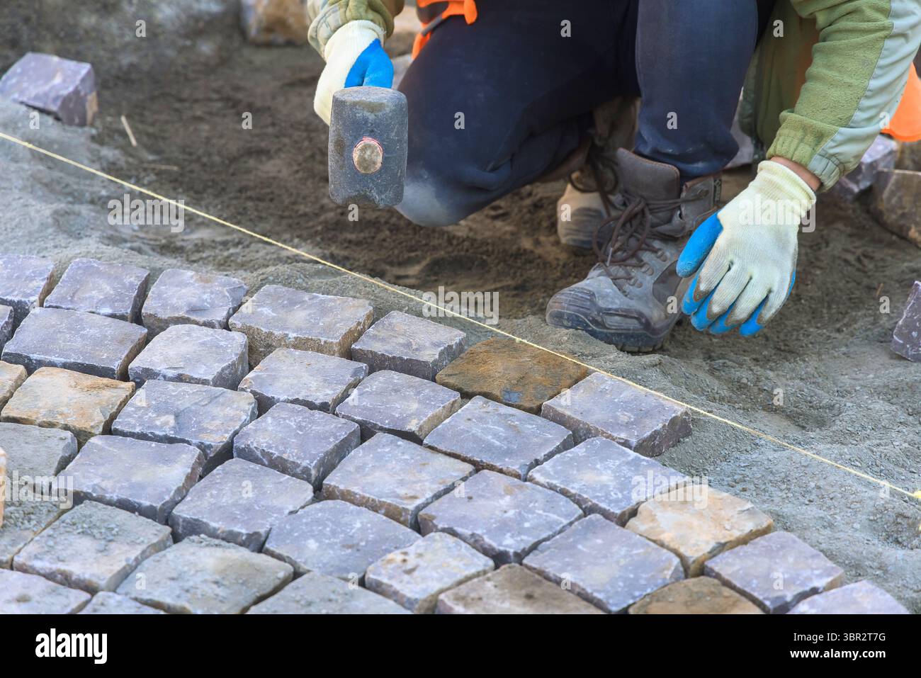 Laborer carefully positions cobblestones while maintaining alignment in construction setting ...