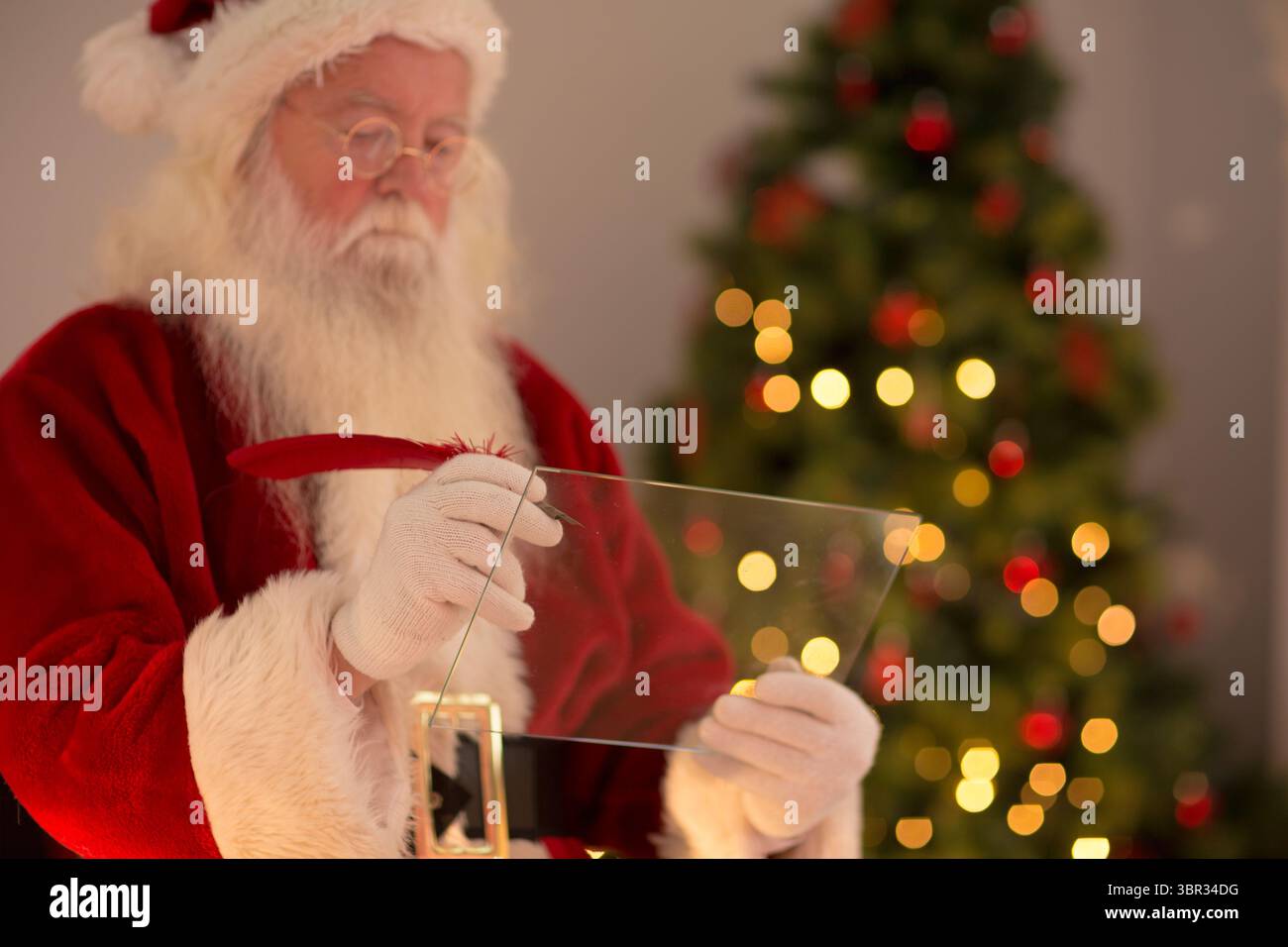 Santa Claus is writing on transparent tablet with red quill by lit tree ...