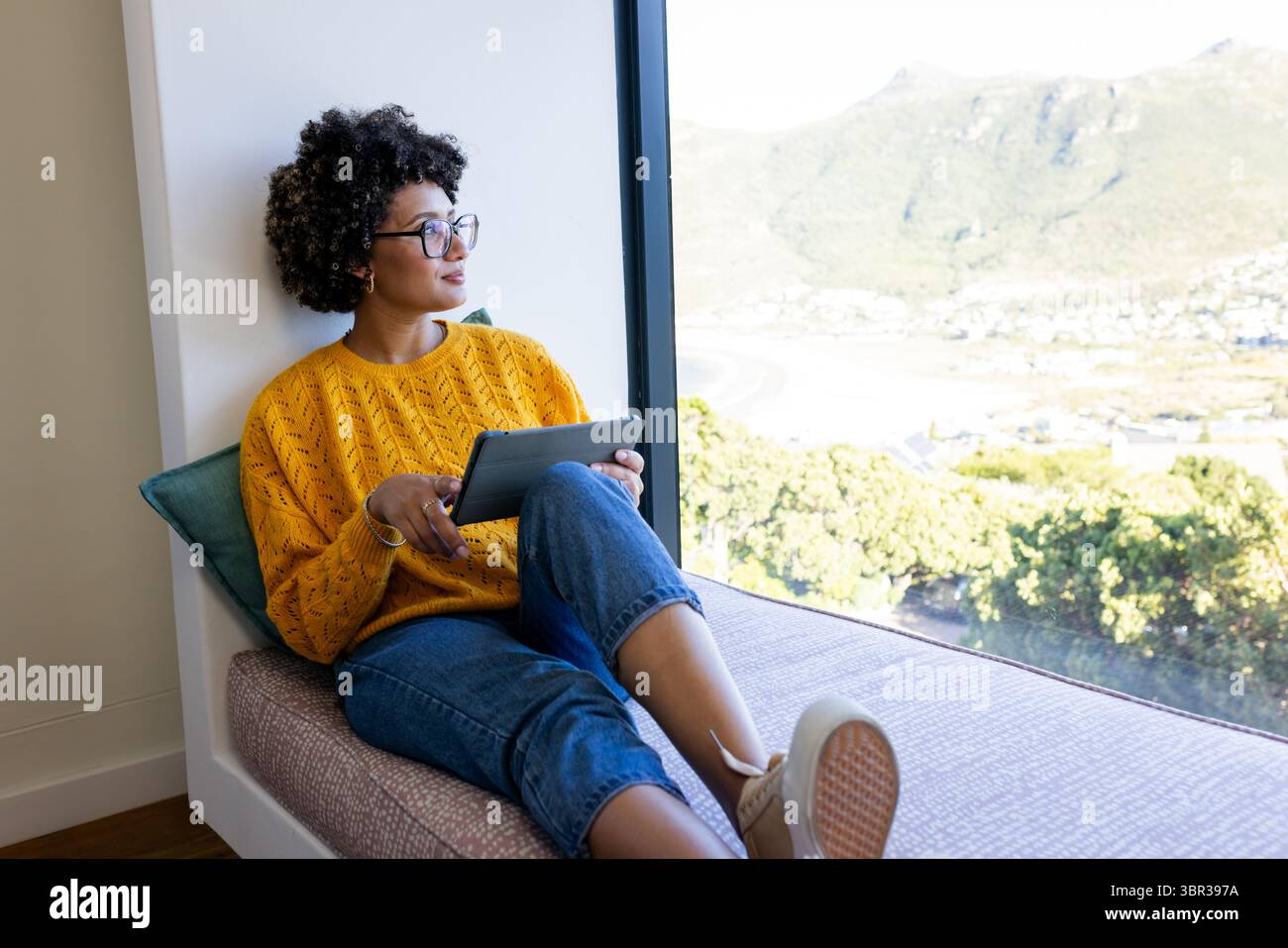 African American woman sitting on window bench holding tablet and ...
