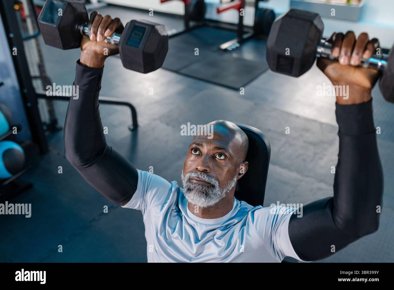 Senior African American man performing overhead dumbbell press on bench ...