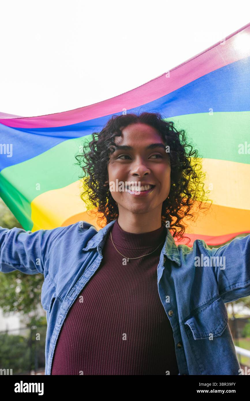 Non-binary person holding large rainbow flag in park smiling wearing denim jacket, maroon top ...