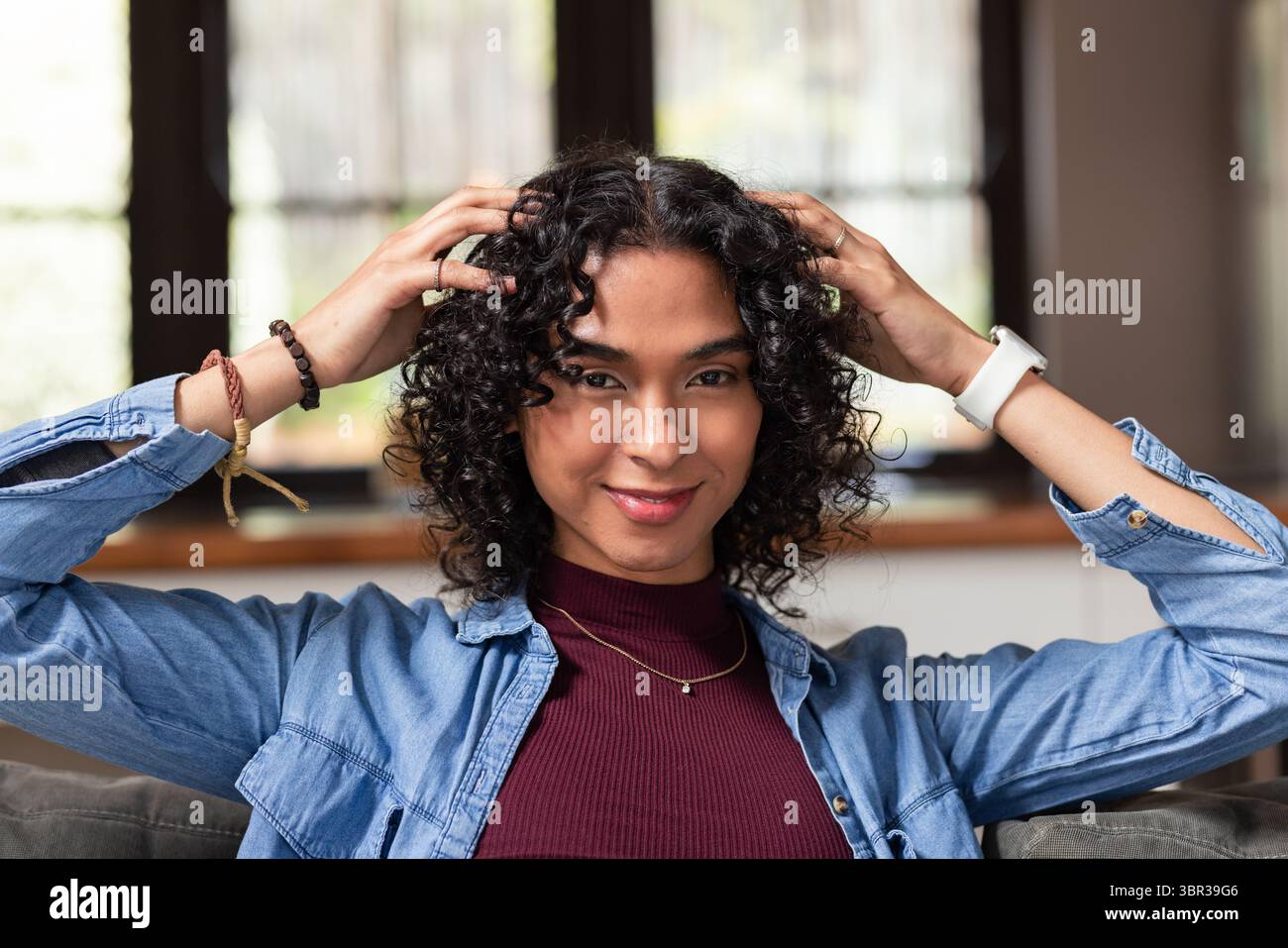 Non-binary person adjusting curly hair on sofa in living room wearing ...