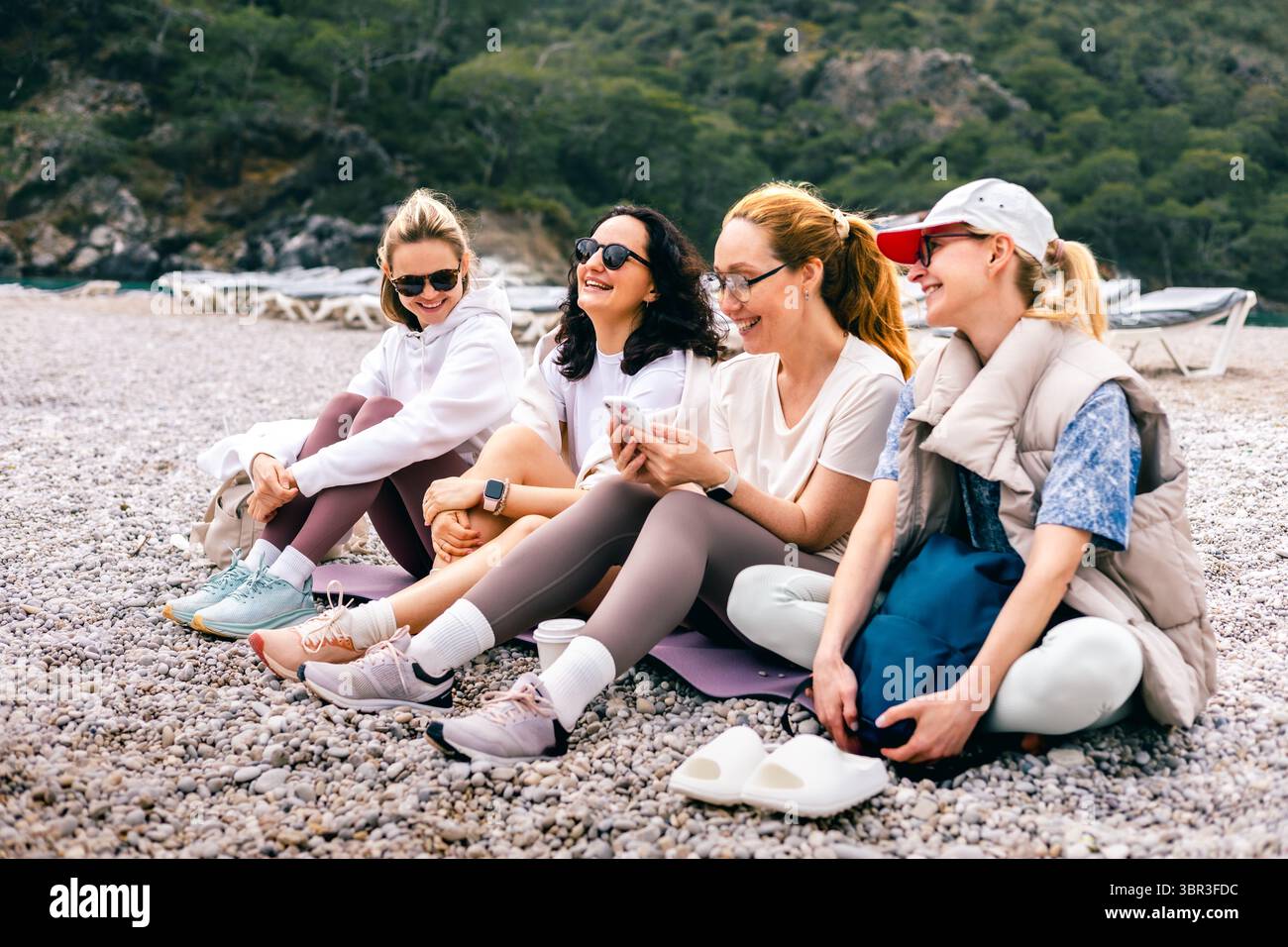 Happy group of women in activewear sitting on beach and laughing ...