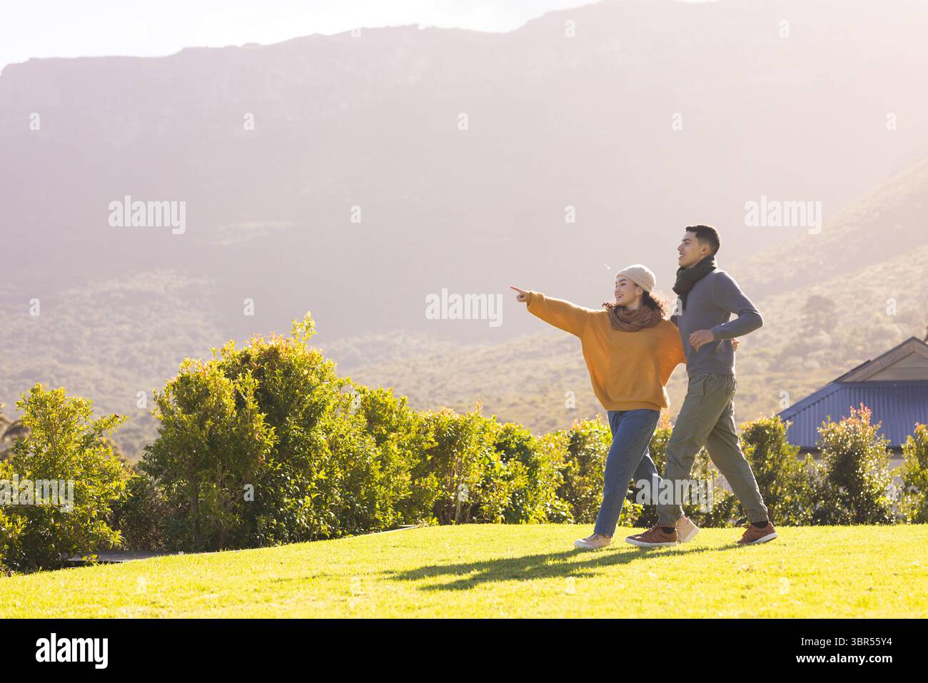 Diverse couple wearing scarves walking on slope near shrubs pointing toward horizon, copy space ...