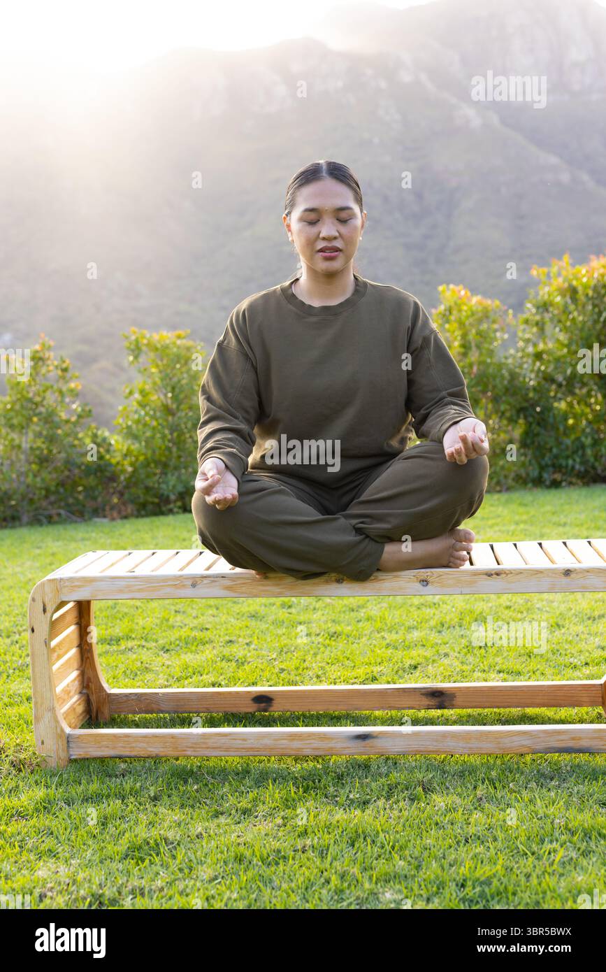 Asian woman meditating cross-legged barefoot on wooden bench in grassy ...