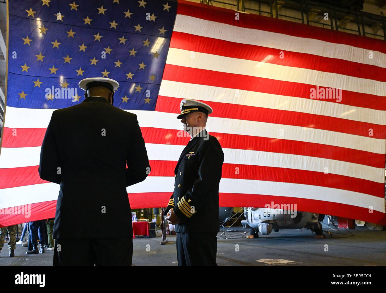 Navy officers are seen standing under a large flag of The United States ...