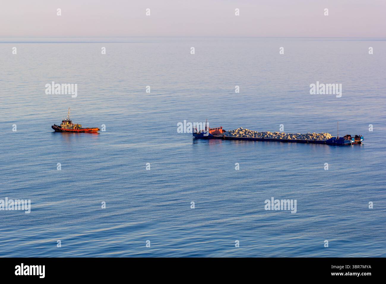A tugboat pulls a large barge loaded with rocks across calm open waters ...
