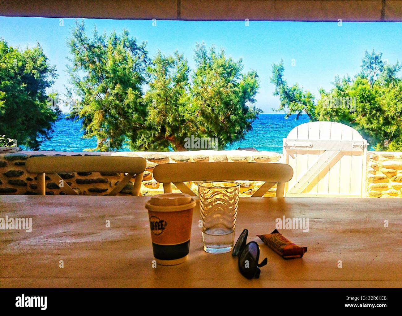 Coffee, water glass, and sunglasses on a shaded terrace table with sea view and trees in Crete ...
