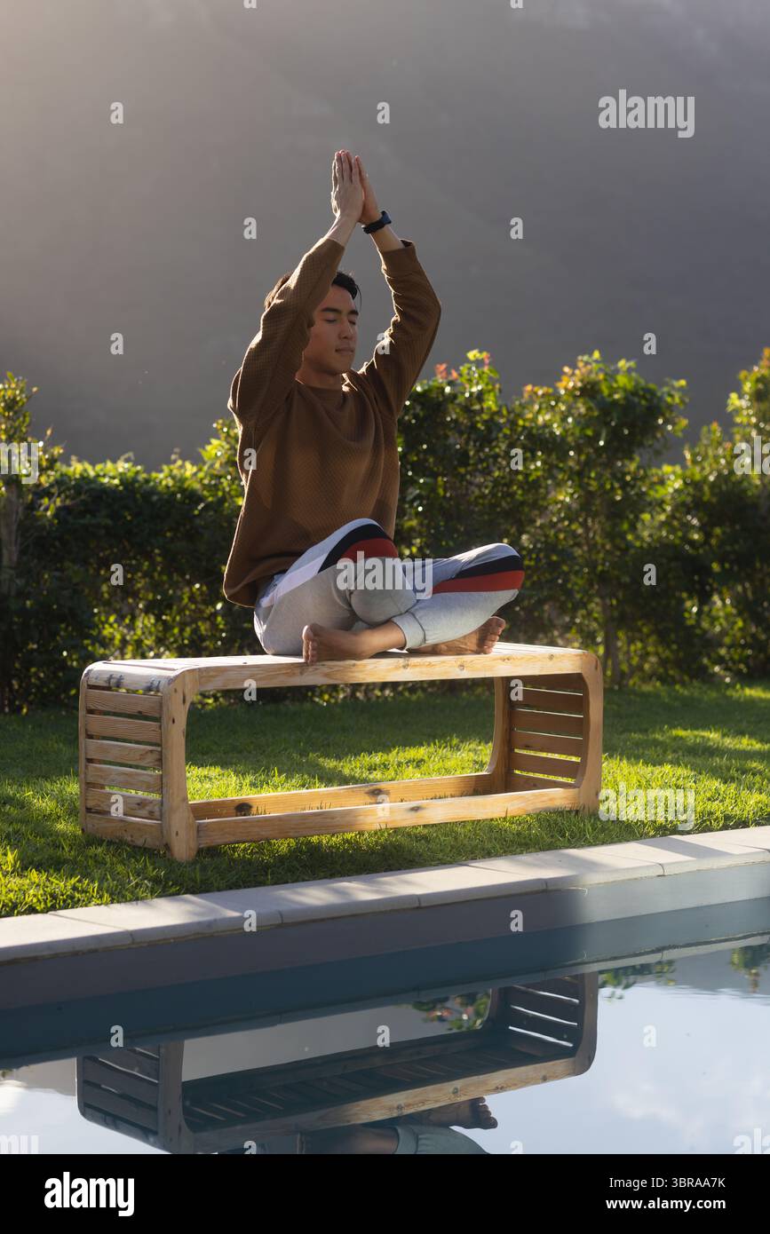 Meditating Asian man sitting on bench on lawn by hedge and pool wearing ...