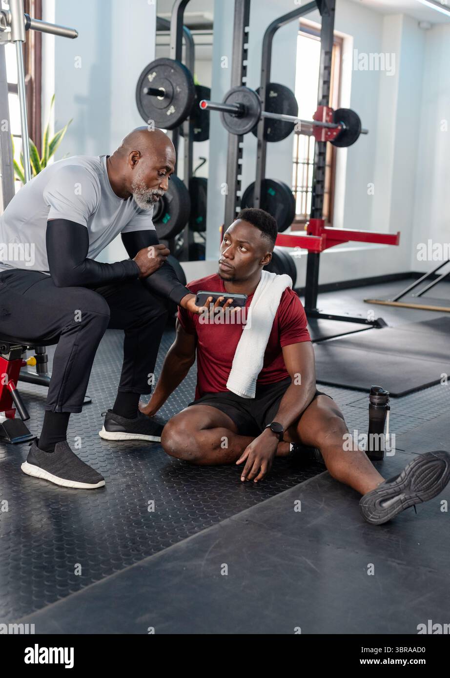 African American trainer and client kneeling beside squat rack in gym ...